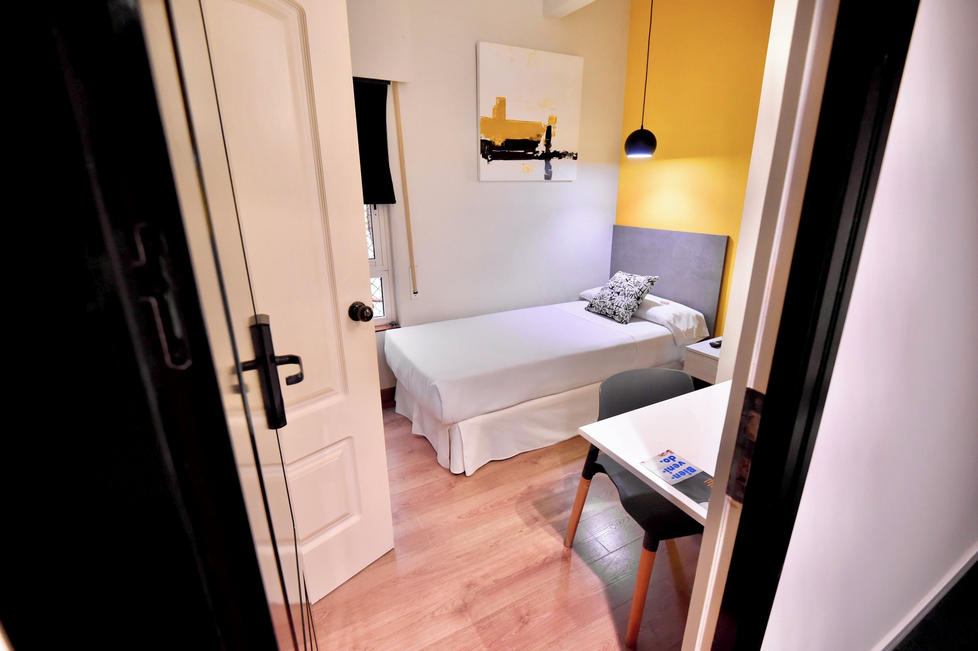 Quarto compacto com uma cama de solteiro, mesa de cabeceira e escrivaninha com cadeira.