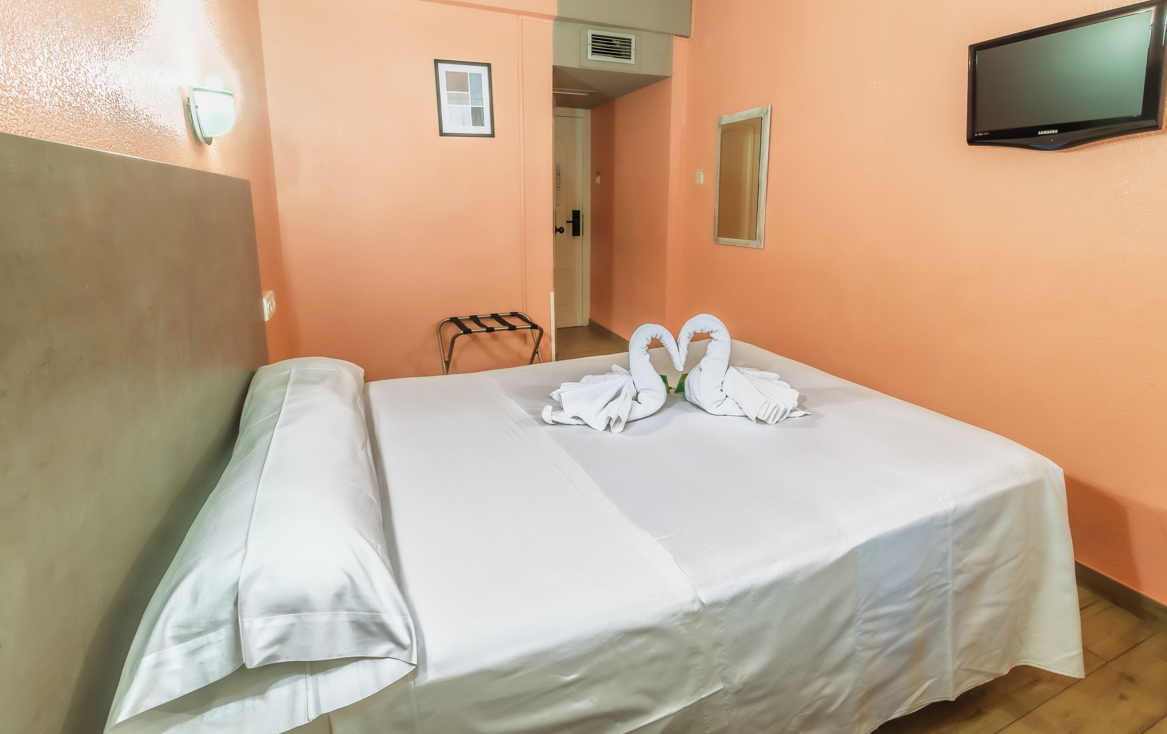 Quarto de hotel com uma cama, televisão presa na parede e corredor que leva a outro quarto.