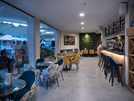 Uma área de bar moderna com cadeiras amarelas, balcão e um espaço de estar com mesas e cadeiras.