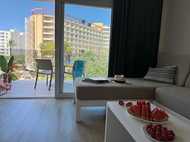 Quarto de hotel moderno com varanda, sofá, mesa de centro com frutas e vista para a cidade.