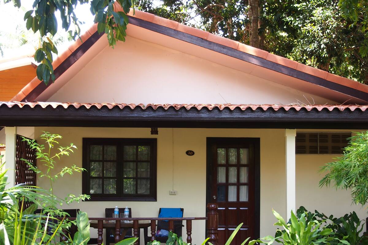 Uma casa com telhado de telhas, varanda com mesa e cadeiras e porta com placa de número.