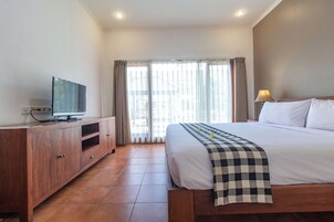 Quarto de hotel com cama, televisão em móvel de madeira, janela com cortinas e abajur no criado-mudo.