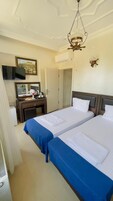 Quarto de hotel com duas camas, uma escrivaninha com cadeira, televisão e ventoinha no teto.