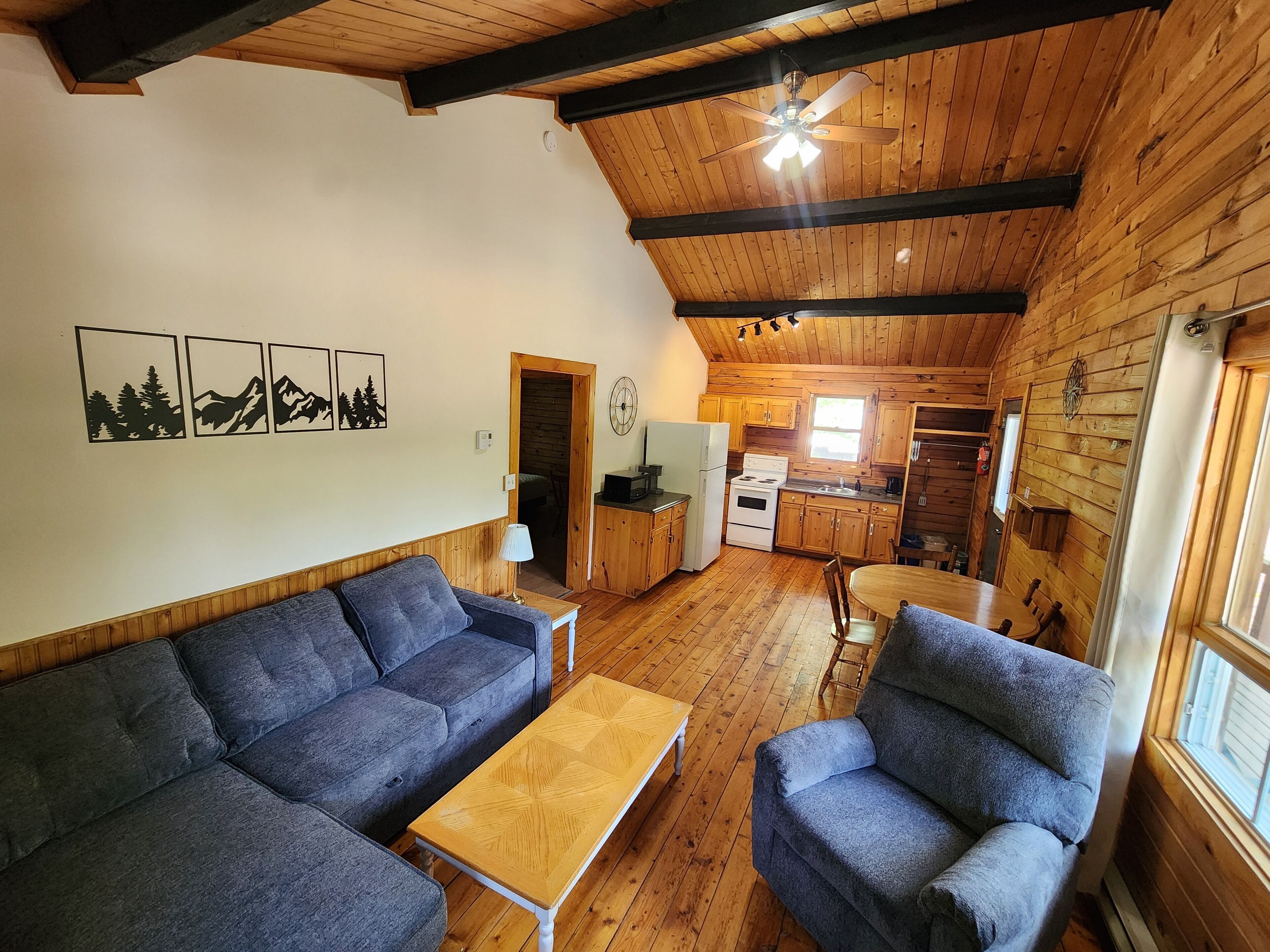 Interior acolhedor de uma cabana, com teto de madeira, um sofá, uma mesa de jantar e uma área de cozinha.