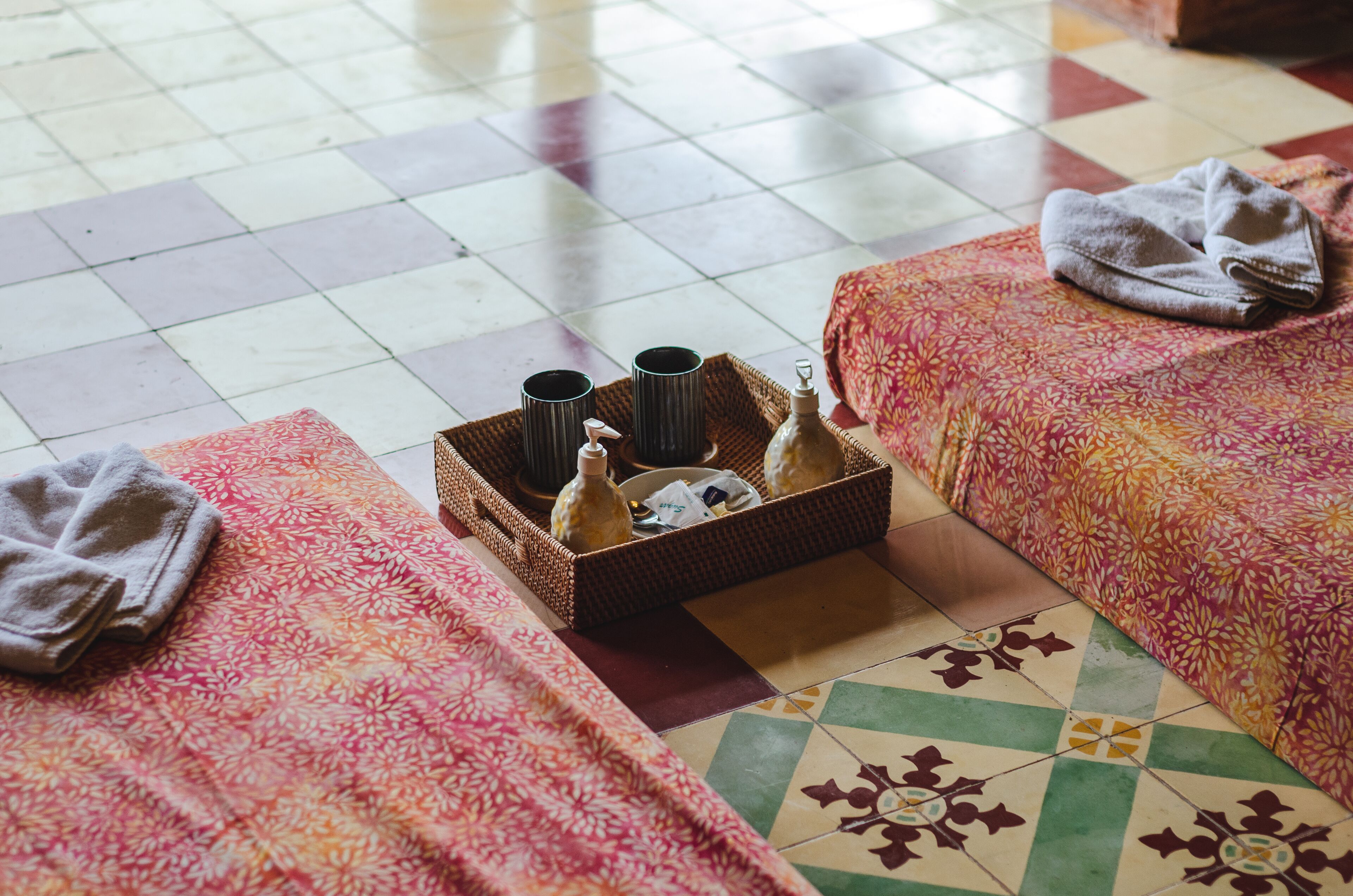 Quarto com colcha estampada, bandeja com xícaras e uma garrafa, e uma toalha no chão.