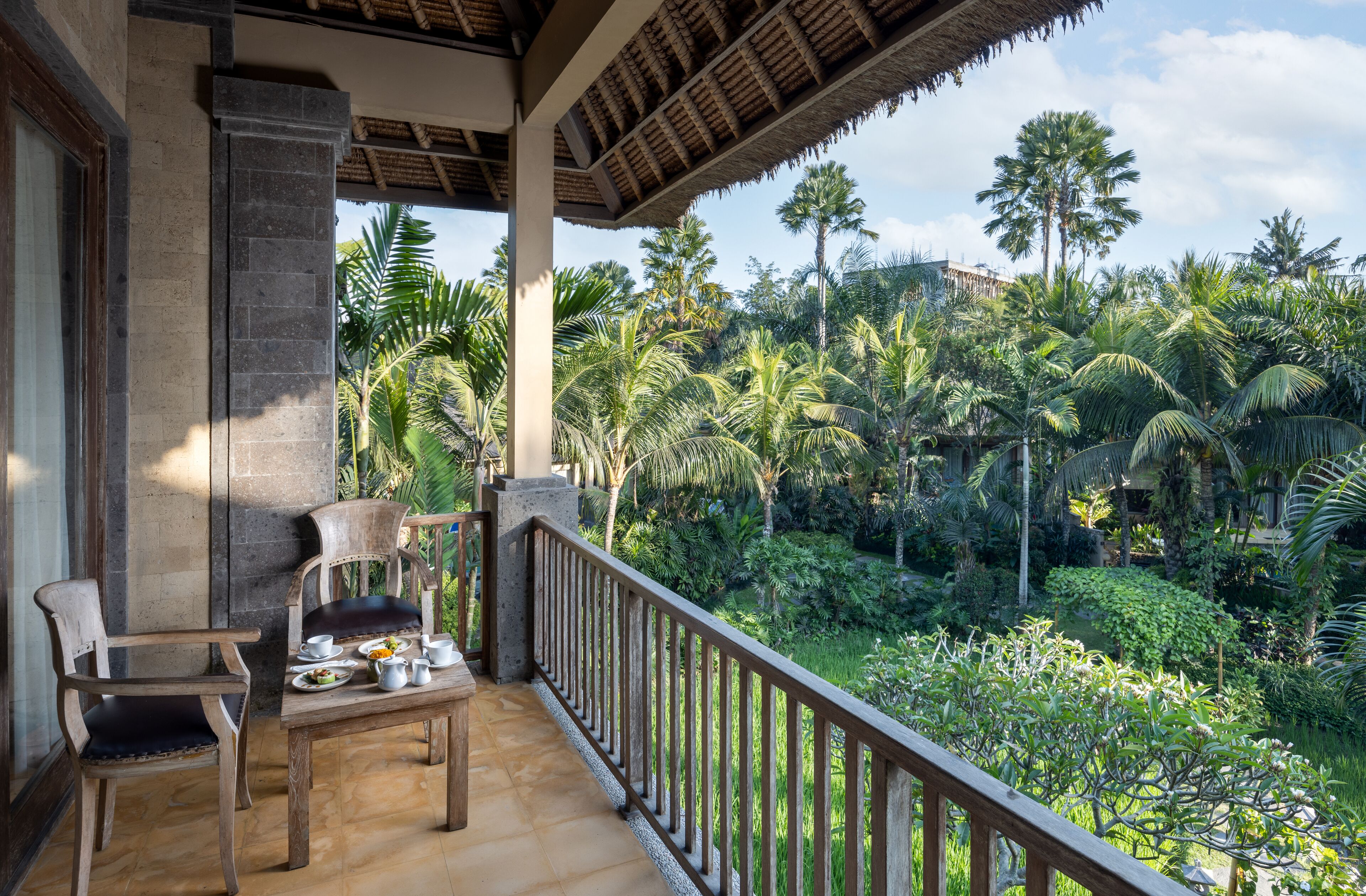 Varanda com mesa posta para refeições, com vista para um jardim exuberante com palmeiras.