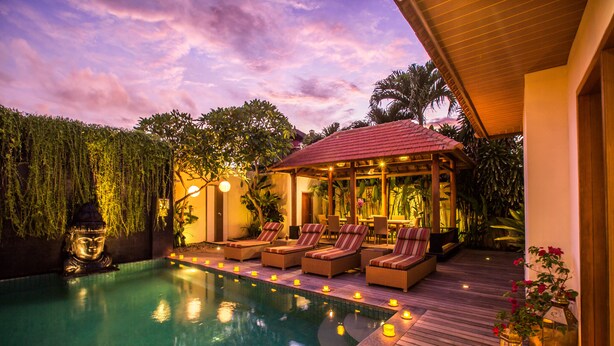Uma área de piscina tranquila com espreguiçadeiras e um espaço coberto para sentar, cercada por vegetação exuberante e um céu ao pôr do sol.