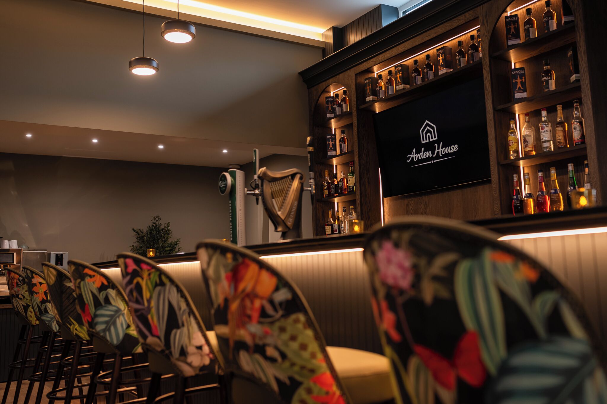 Uma área de bar com um letreiro que diz 'Alden House', banquetas e prateleiras com garrafas.
