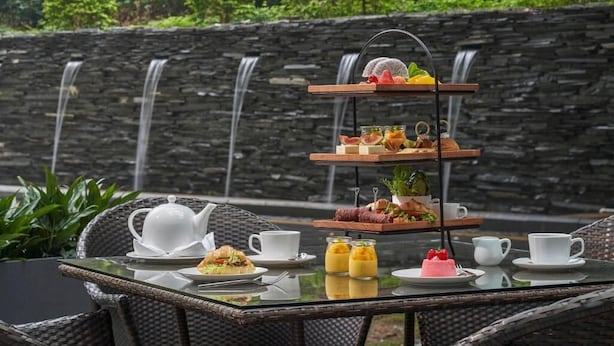 Uma bandeja escalonada com frutas e doces, um bule de chá, xícaras e um copo de suco sobre uma mesa ao ar livre, com uma parede de pedra e uma cascata ao fundo.