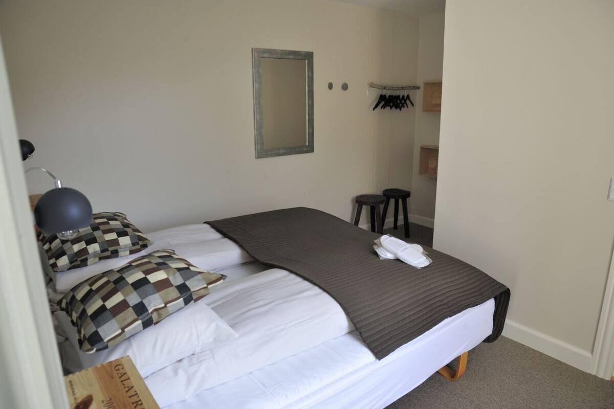 Cama bem arrumada com travesseiro xadrez, cobertor escuro e toalha branca. Há um banquinho e um espelho na parede.