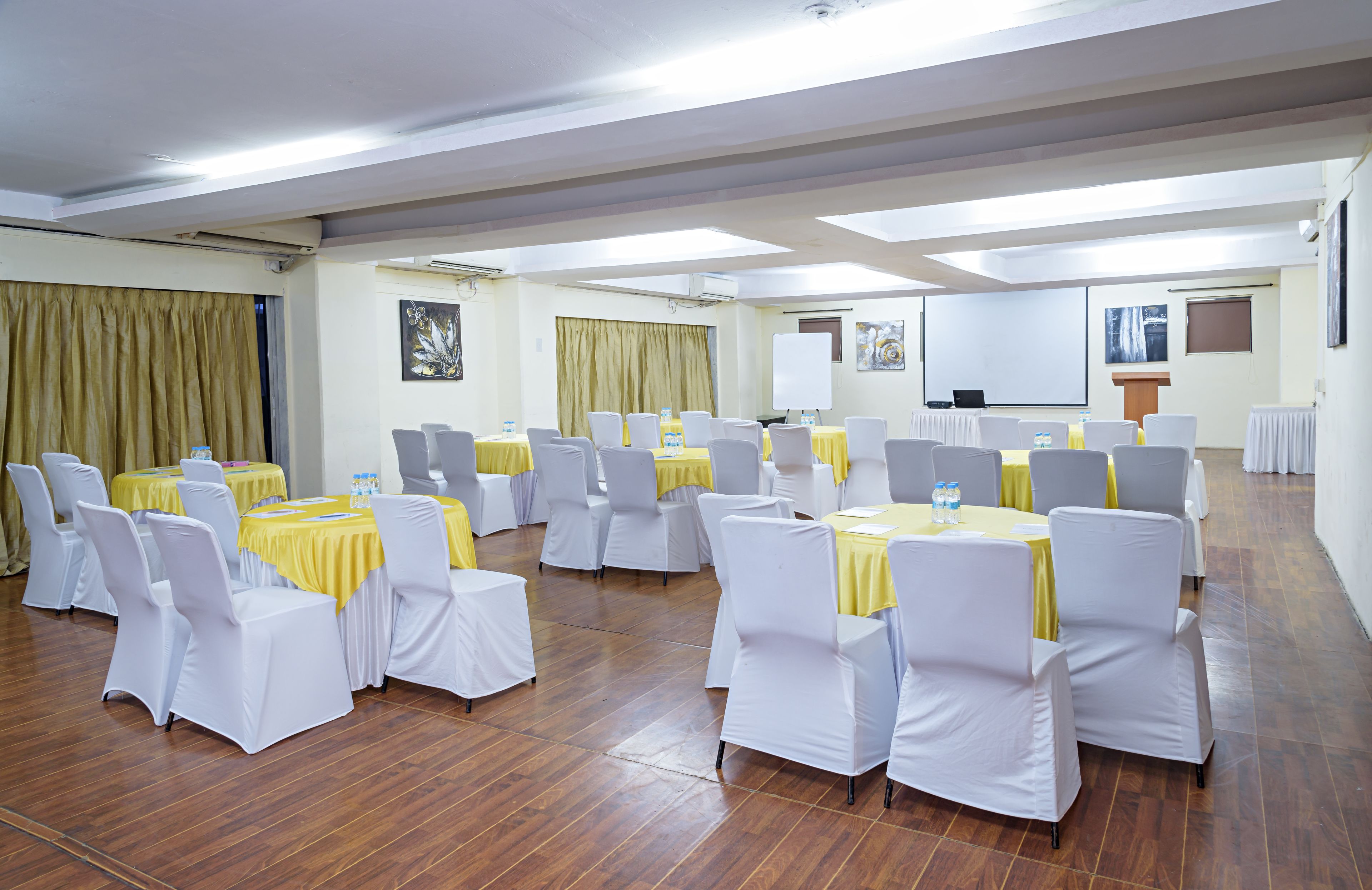 Sala de conferências com várias mesas e cadeiras, unidades de ar condicionado e uma tela de projeção.