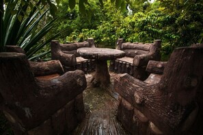 Uma área de estar ao ar livre rústica, com bancos de madeira e uma mesa central, rodeada por vegetação exuberante.
