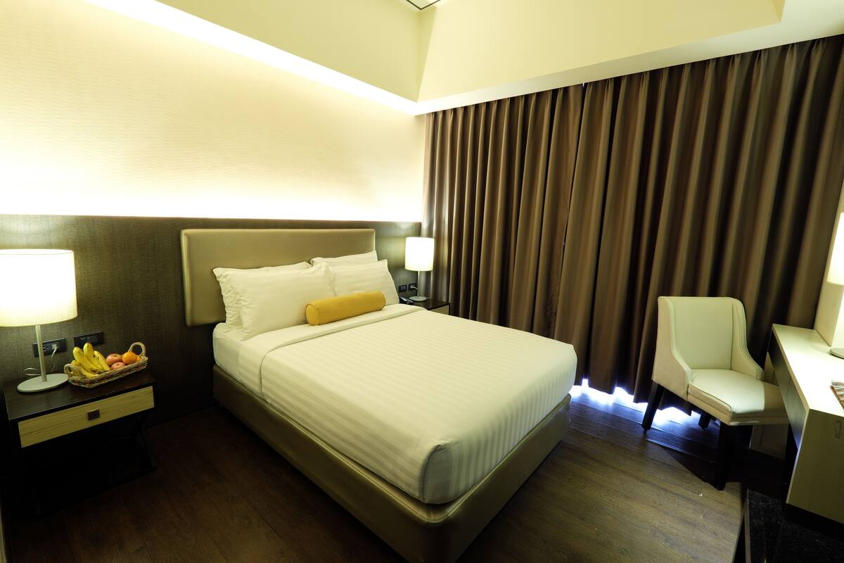 Quarto de hotel com uma cama grande, uma escrivaninha, uma cadeira, uma mesa de cabeceira com abajur e uma cesta de frutas.