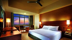 Quarto de hotel com uma cama grande, uma escrivaninha, uma cadeira, abajur, mesa de cabeceira com telefone e janela com vista para o pôr do sol.