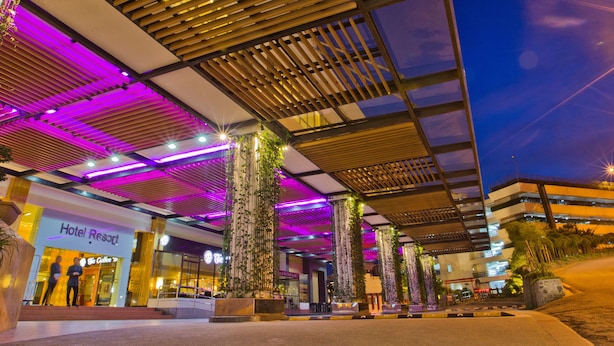 Entrada de hotel com design moderno, destacando fachada de vidro, área externa coberta com ripas de madeira e plantas pendentes, e um letreiro visível de cafeteria.