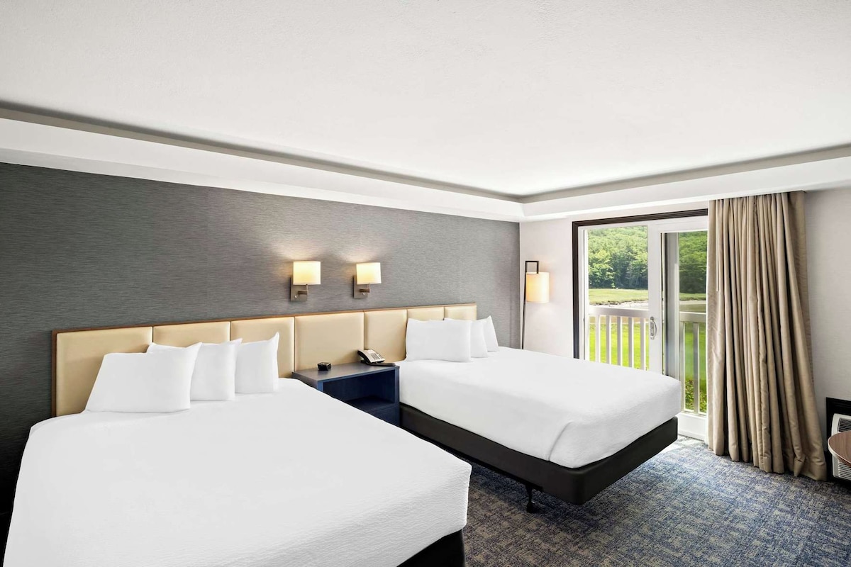Quarto de hotel com duas camas, mesa de cabeceira, luminária e varanda com cortinas.