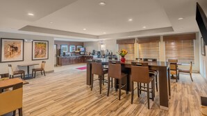 Um hall de hotel espaçoso com área de jantar central, um bar e uma cozinha ao fundo.