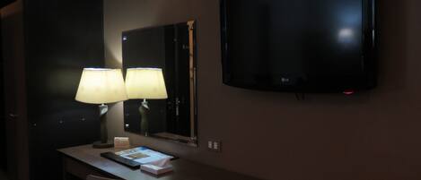Quarto de hotel com televisão de tela plana presa na parede, uma escrivaninha com luminária e uma cadeira.
