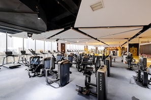 Academia moderna com diversos equipamentos de exercício, grandes janelas e vista para a paisagem urbana.