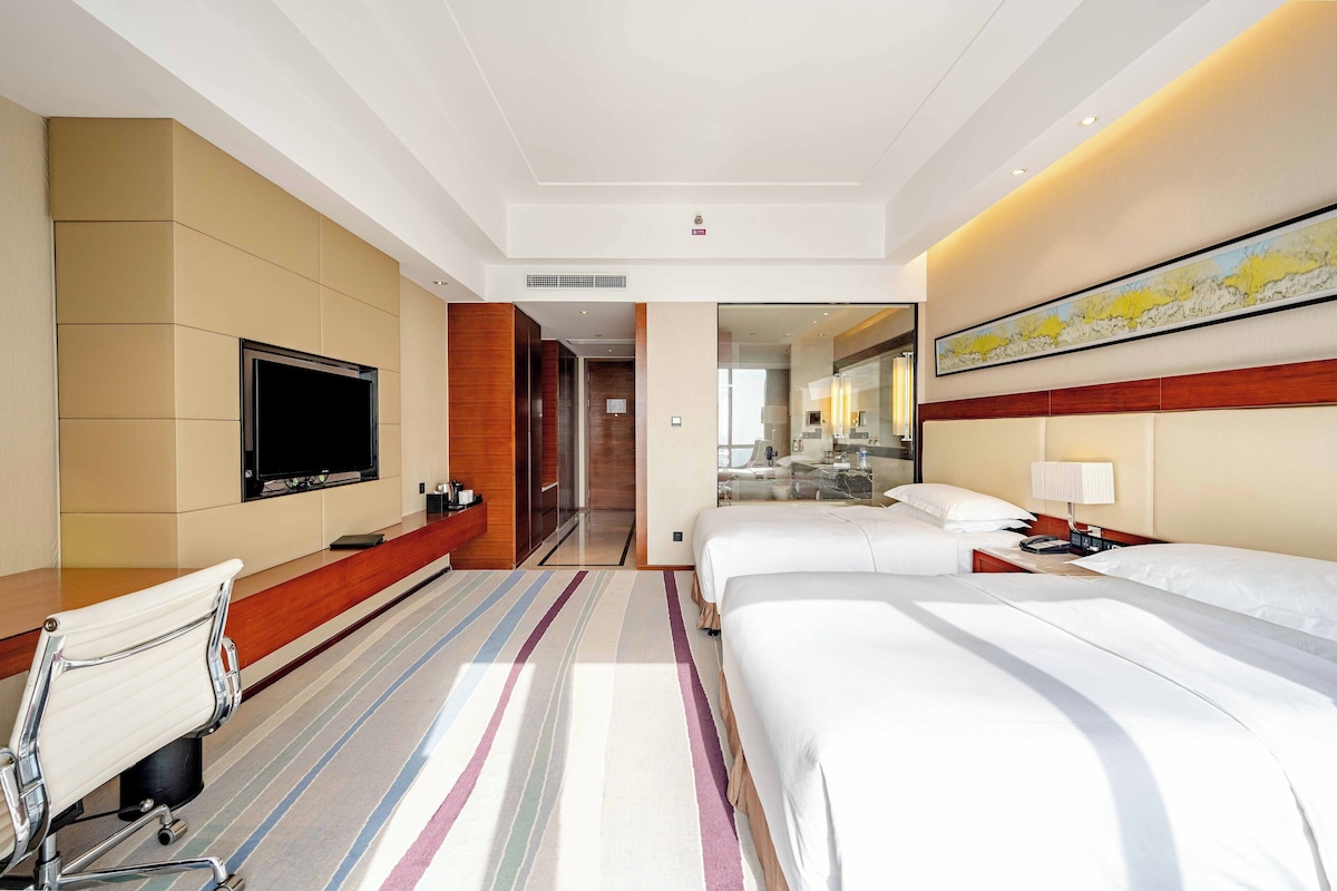Quarto de hotel com televisão, duas camas, uma escrivaninha e um banheiro visível por uma porta aberta.