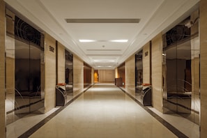 Corredor moderno de hotel com elevadores, piso de mármore e iluminação embutida.