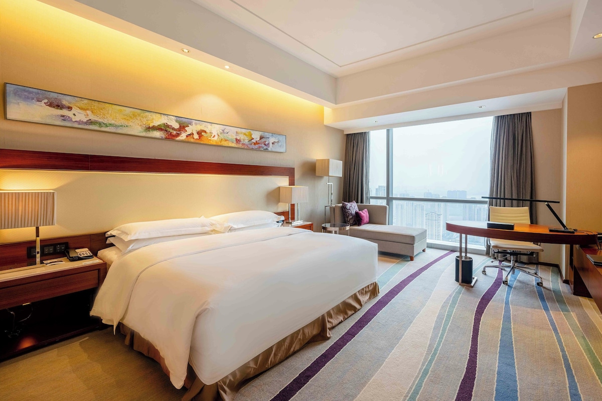 Quarto de hotel com cama grande, escrivaninha, cadeira, sofá, janela com vista e um quadro na parede.