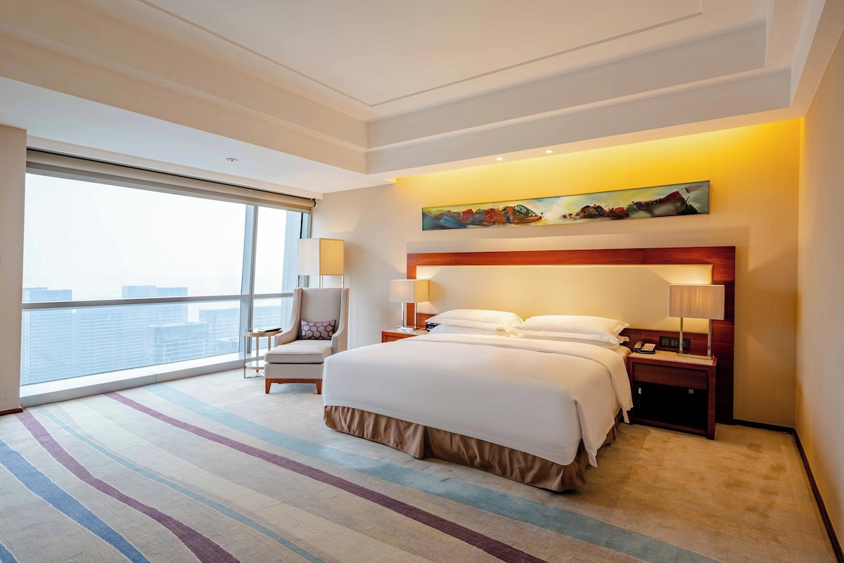 Quarto de hotel com uma cama grande, uma mesa de cabeceira com abajur, uma cadeira e uma janela ampla com vista para a cidade.