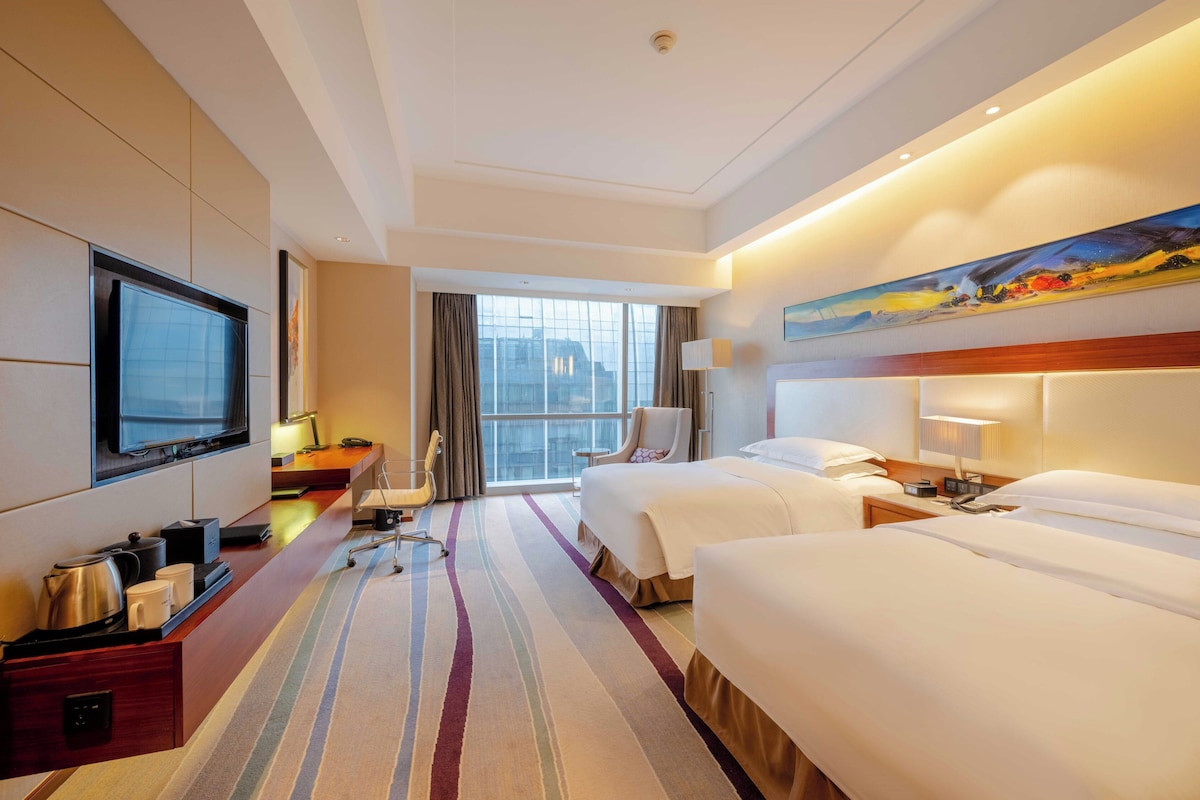 Quarto de hotel com duas camas, televisão, secretária com cafeteira e uma janela grande.