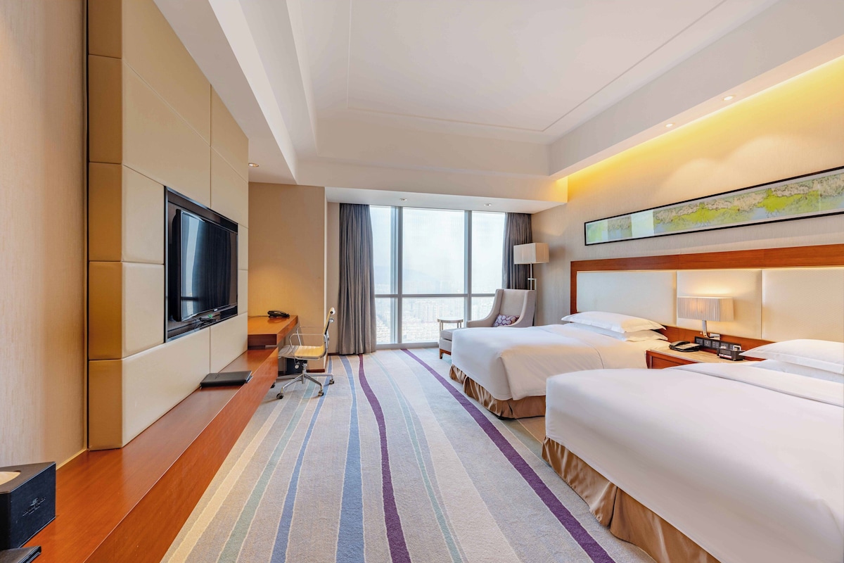 Quarto de hotel com duas camas, televisão, secretária e uma janela grande.