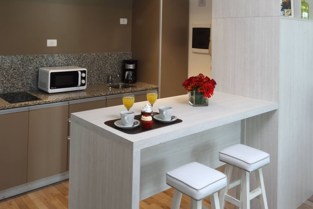 Uma cozinha moderna com uma ilha central, banquetas brancas, micro-ondas, cafeteira e um vaso com flores.