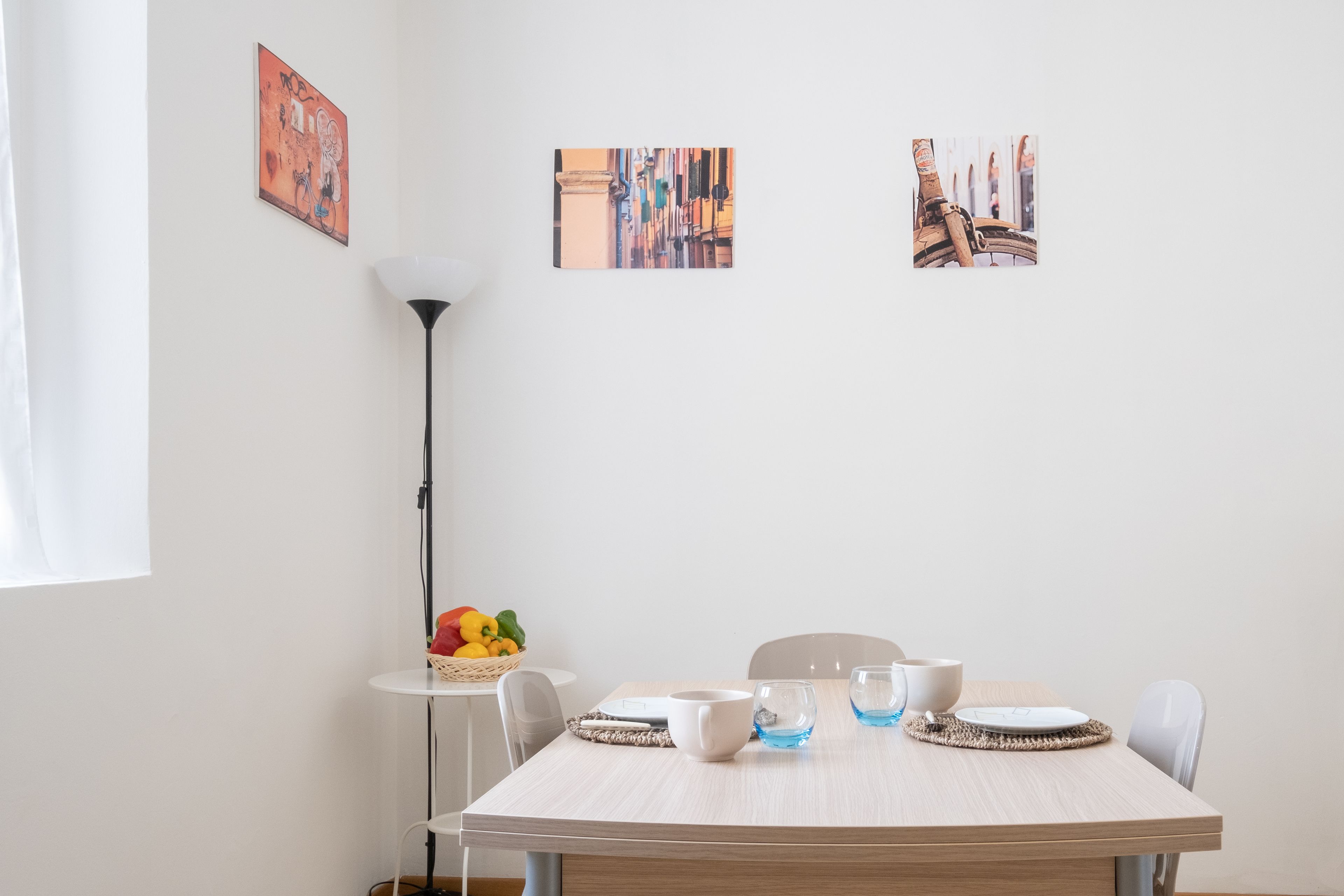 Uma área de refeições com uma mesa posta para dois, uma cesta de frutas e duas molduras na parede.