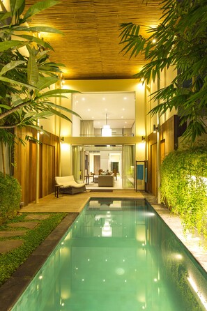 Uma área interna de piscina moderna com piscina transparente, deck de madeira e vegetação ao redor.