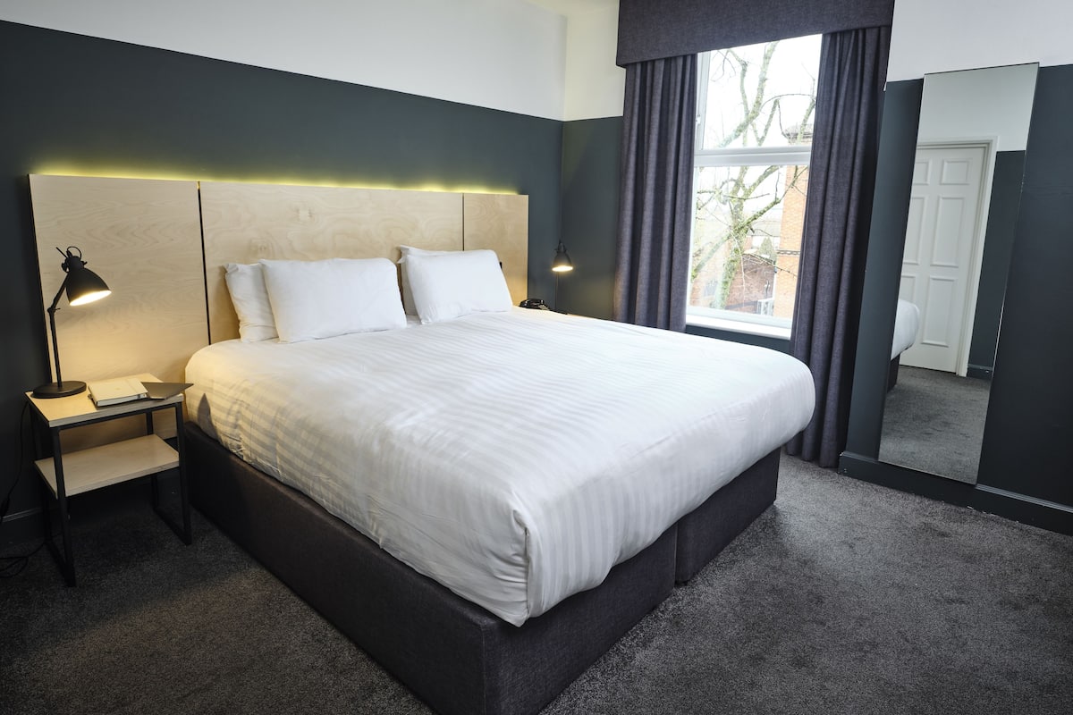 Quarto de hotel moderno com uma cama grande, luminárias de cabeceira, mesa de apoio e janela com cortinas.
