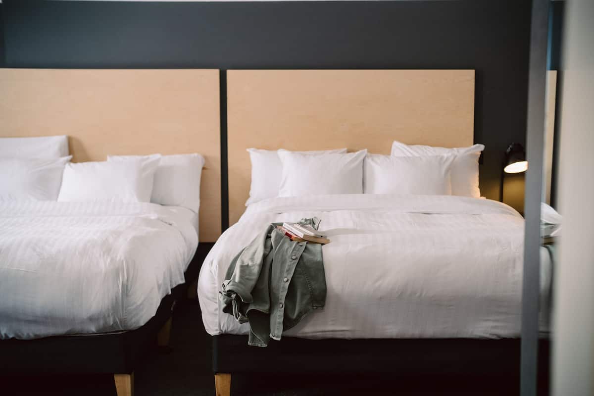 Duas camas com roupa de cama branca e cabeceiras de madeira, um par de calças de ganga e alguns livros sobre uma das camas.