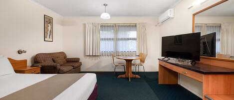 Quarto de hotel com uma cama, uma escrivaninha com televisão, uma cadeira, uma mesa e uma janela com cortinas.