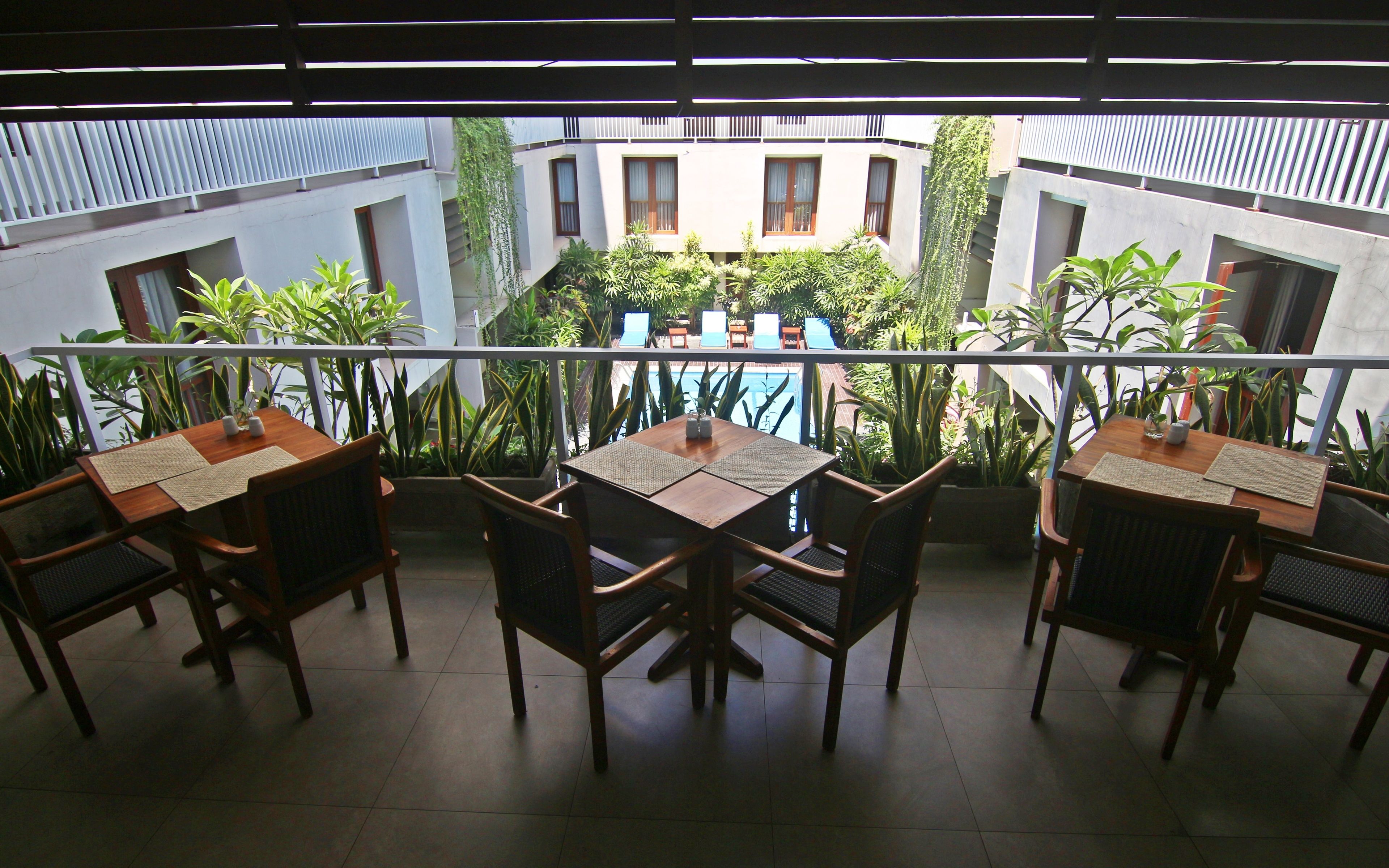 Um terraço com mesas e cadeiras de madeira, rodeado por plantas em vasos e com vista para uma piscina.