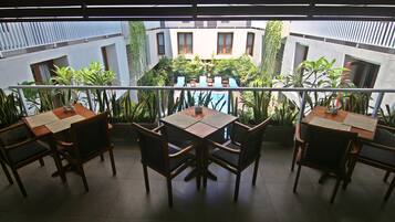 Um terraço com mesas e cadeiras de madeira, rodeado por plantas em vasos e com vista para uma piscina.