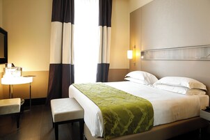 Quarto de hotel com cama, mesa de cabeceira, luminária e janela com cortinas.