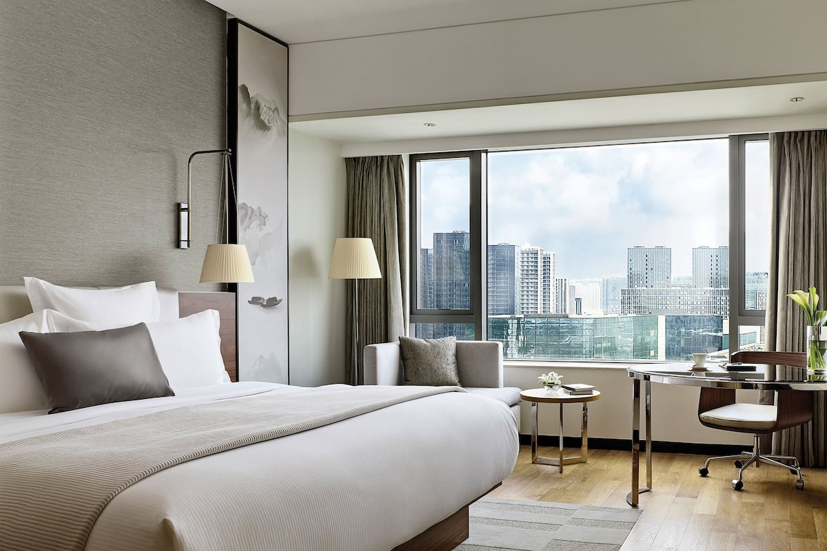 Quarto de hotel moderno com uma cama grande, uma escrivaninha e vista para o horizonte da cidade.