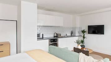 Uma cozinha moderna com armários brancos, micro-ondas, torradeira e chaleira. Há uma TV montada na parede e uma planta no balcão.
