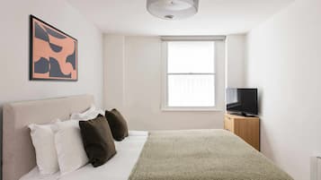 Quarto com cama, travesseiros, cobertor, televisão sobre suporte de madeira, janela e luminária no teto.