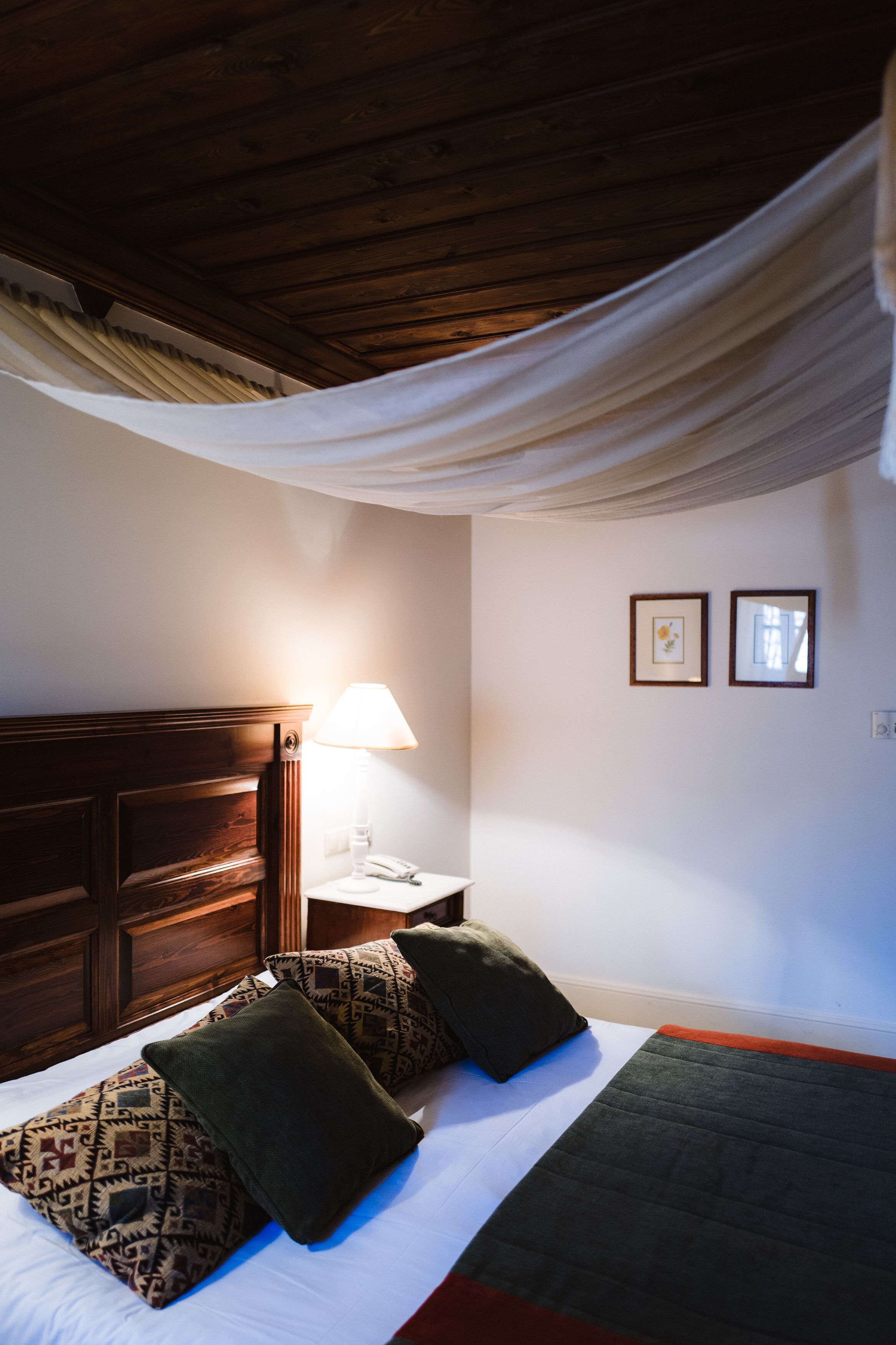 Quarto com cabeceira de madeira, luminária de cabeceira, almofadas estampadas e cobre-leito.
