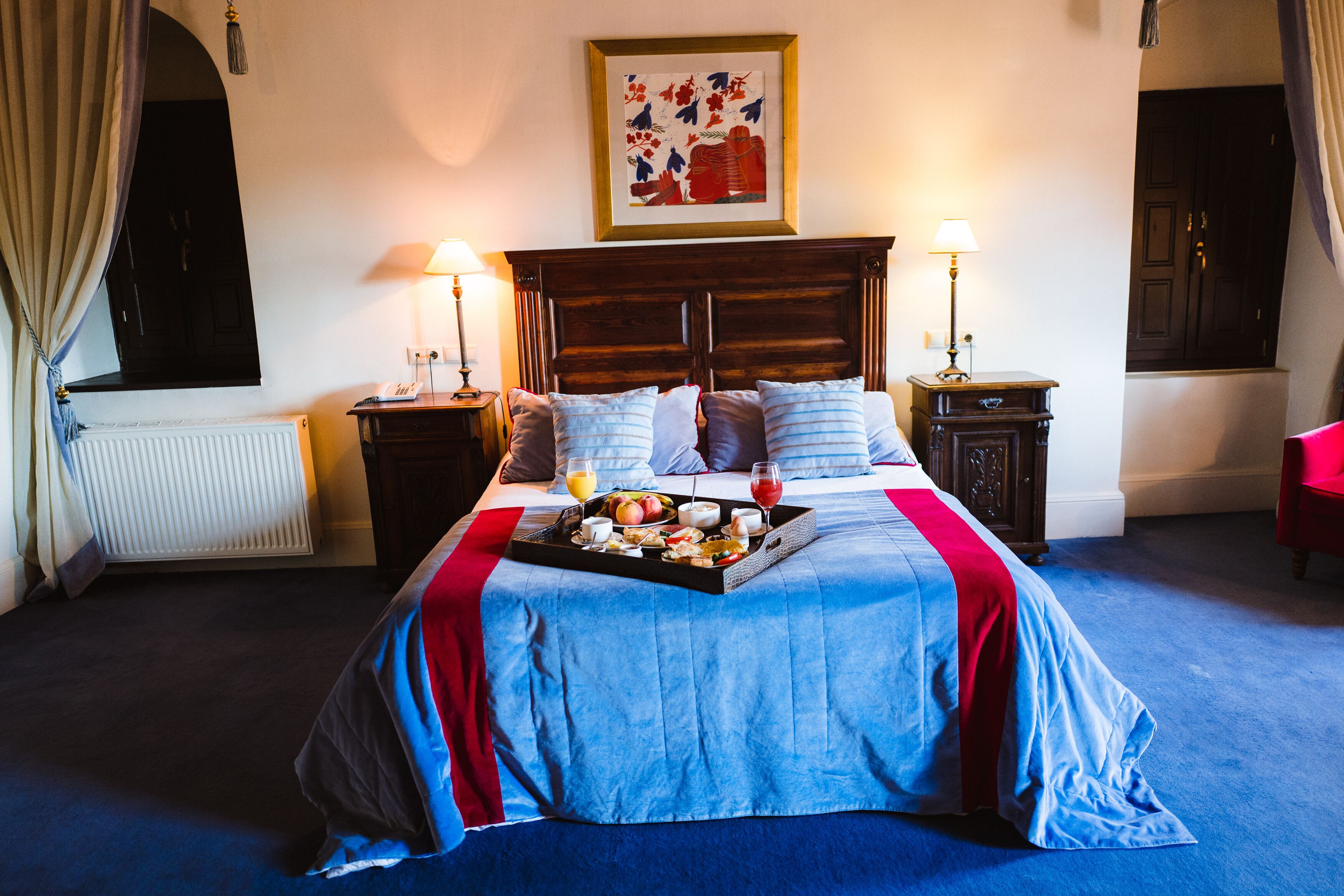 Quarto de hotel com uma cama grande, duas mesas de cabeceira, uma obra de arte emoldurada sobre a cama, um radiador e uma janela com cortinas.