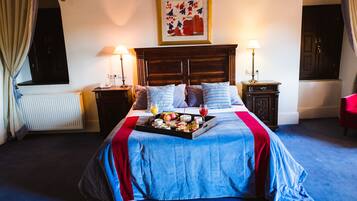 Quarto de hotel com uma cama grande, duas mesas de cabeceira, uma obra de arte emoldurada sobre a cama, um radiador e uma janela com cortinas.