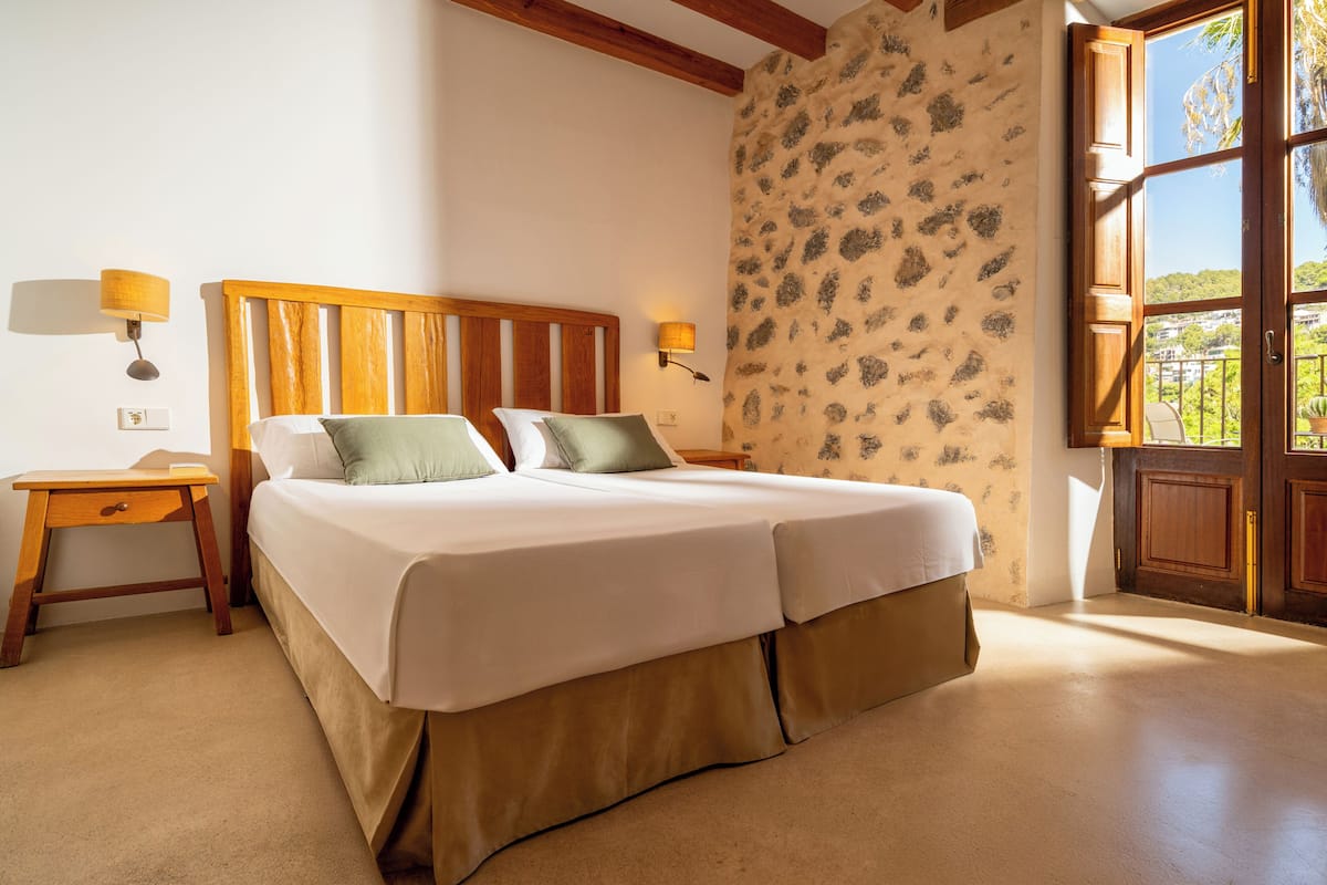 Uma cama de casal com cabeceira de madeira, duas luminárias de apoio, um criado-mudo e vista para uma parede de pedra pela janela.