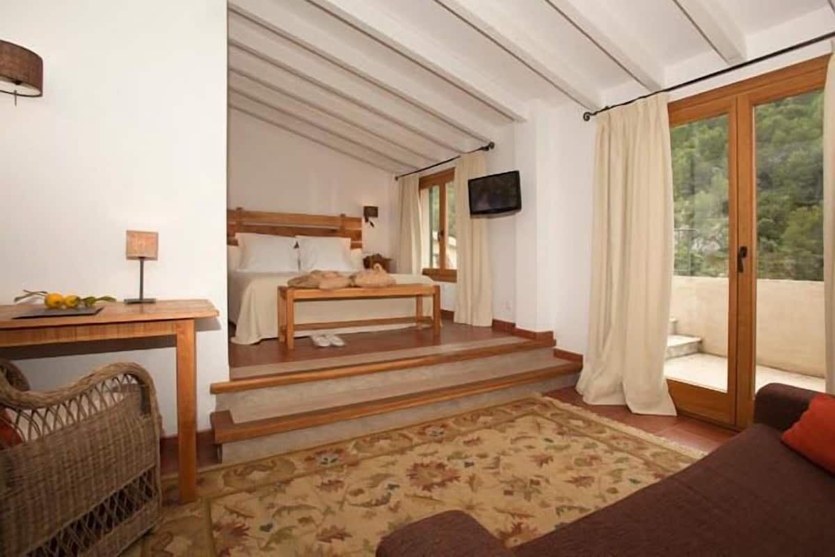 Quarto com cama de madeira, uma secretária pequena, televisão e vista para o exterior através de uma porta de vidro.