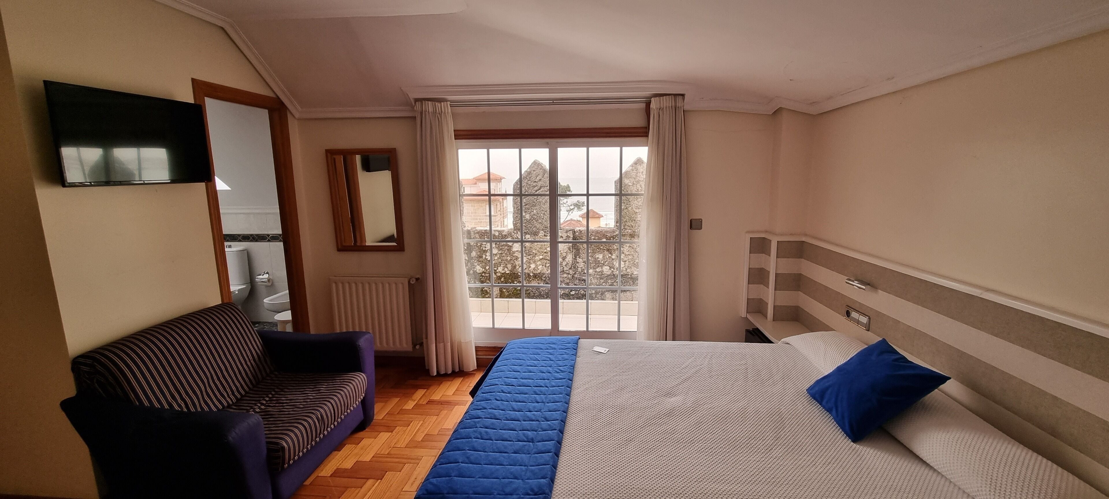 Quarto de hotel com cama, sofá, televisão, janela com cortinas e vista para prédios.