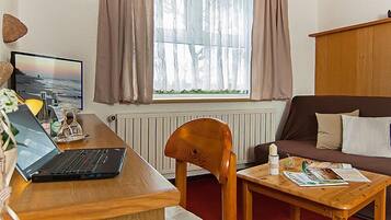 Quarto com escrivaninha de madeira, laptop, cadeira, mesa de centro de madeira, sofá e janela com cortinas.
