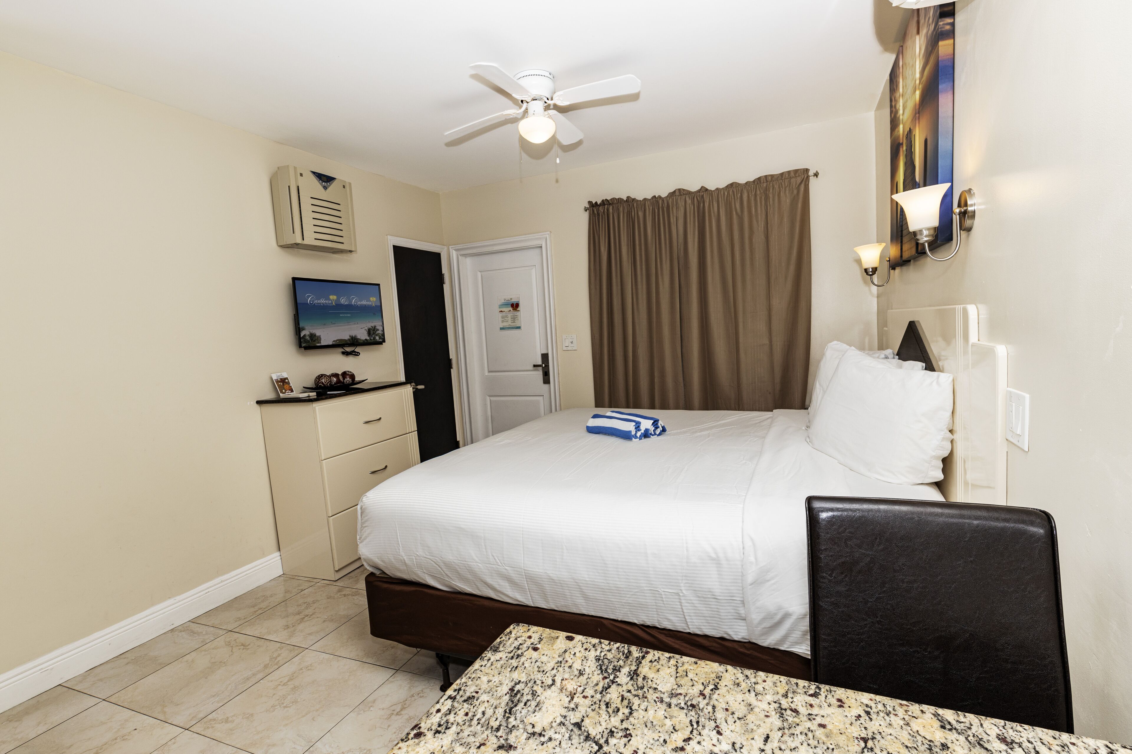 Quarto de hotel com cama, ventoinha no teto, televisão, escrivaninha e janela com cortinas.