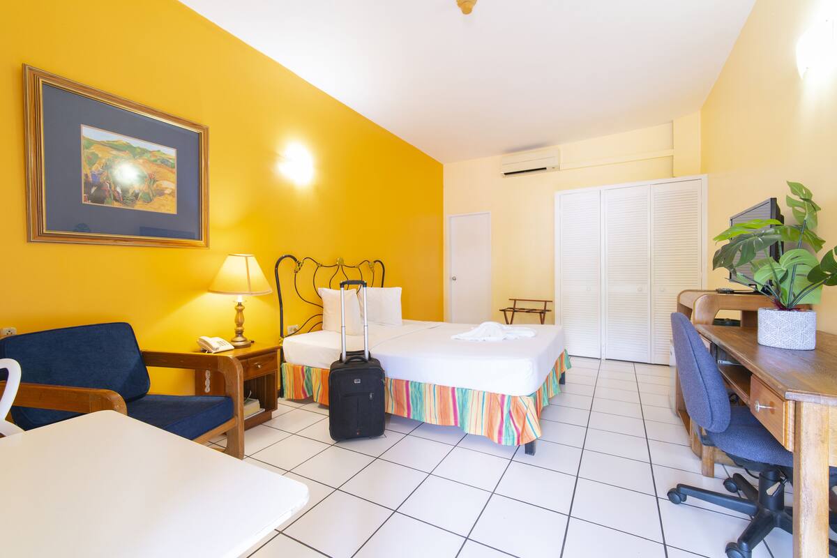 Quarto de hotel com cama, escrivaninha, cadeira, mala, abajur e um quadro na parede.